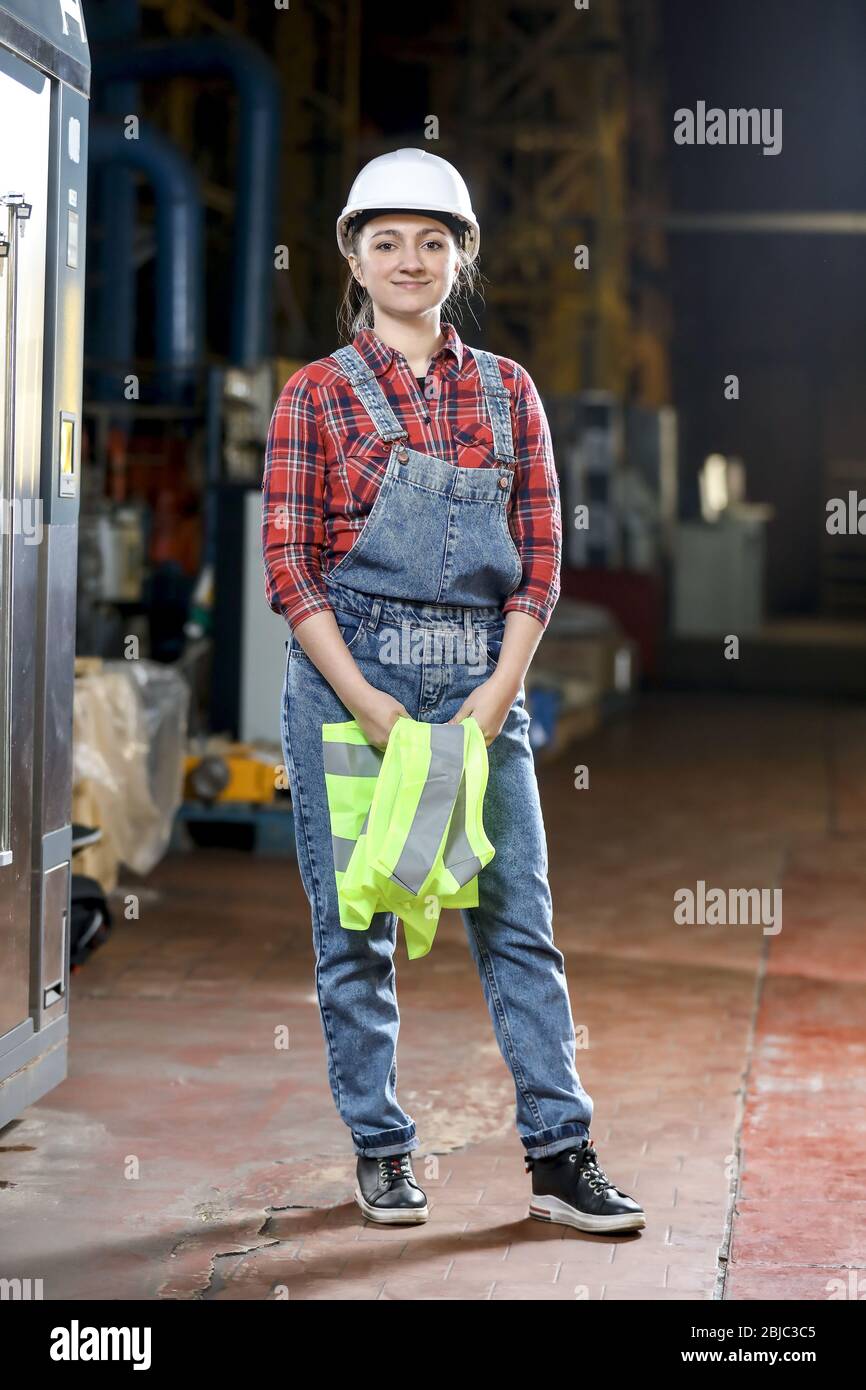 Young girl in a work dress and white hard hat in a factory. Woman in a ...