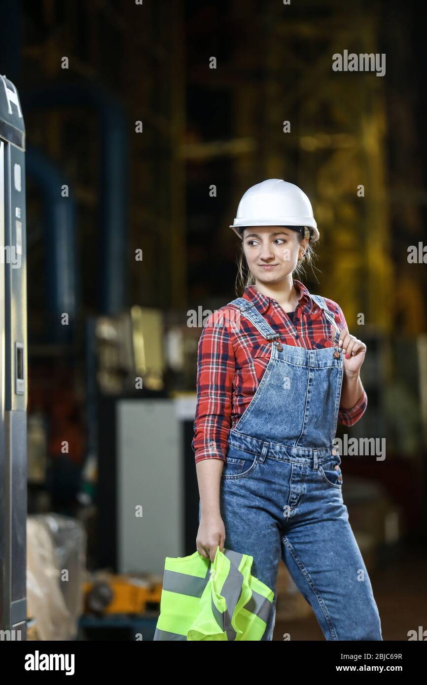 Young girl in a work dress and white hard hat in a factory. Woman in a ...