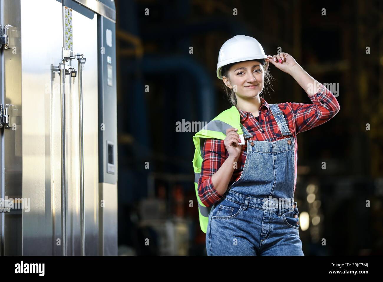 Young girl in a work dress and white hard hat in a factory. Woman in a ...