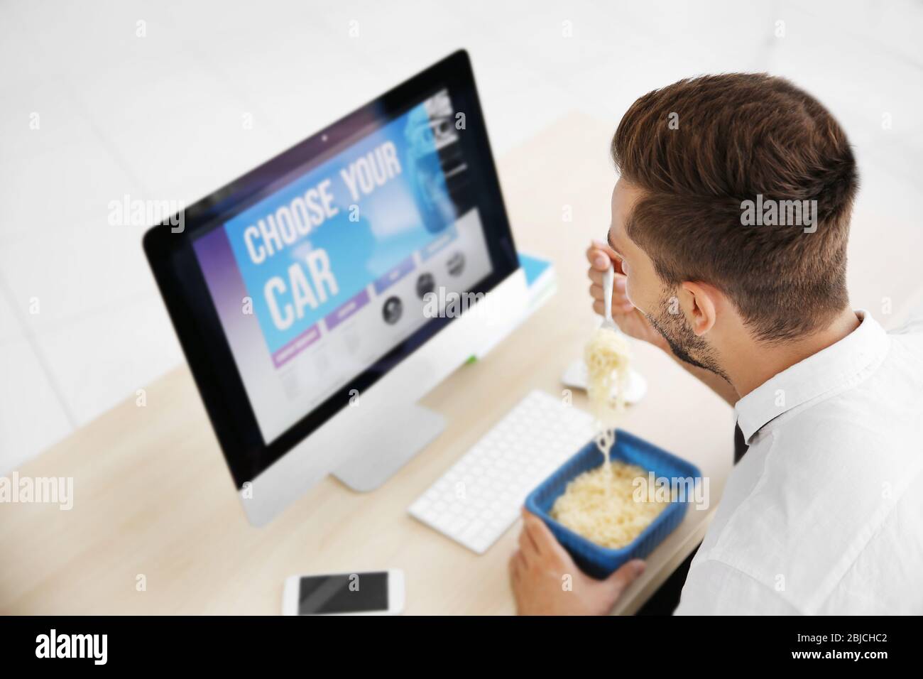 Young man eating instant noodles while working with computer in office ...