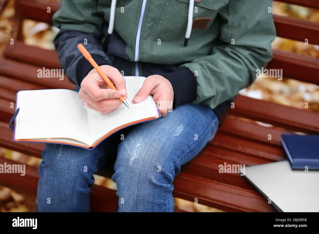 Teenager writing in notebook while sitting on bench in autumn park ...