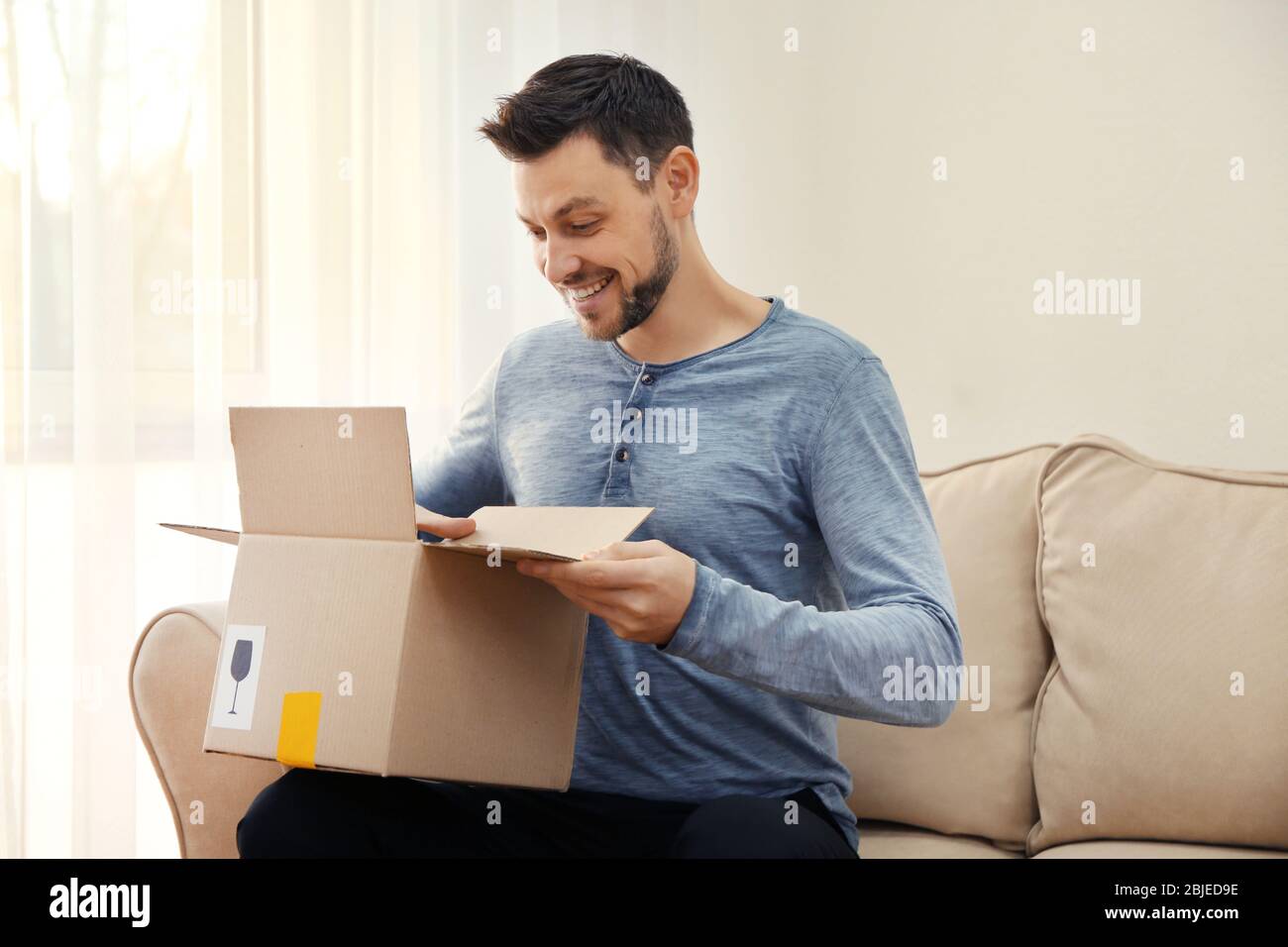 Handsome young man opening box with parcel while sitting on sofa at ...
