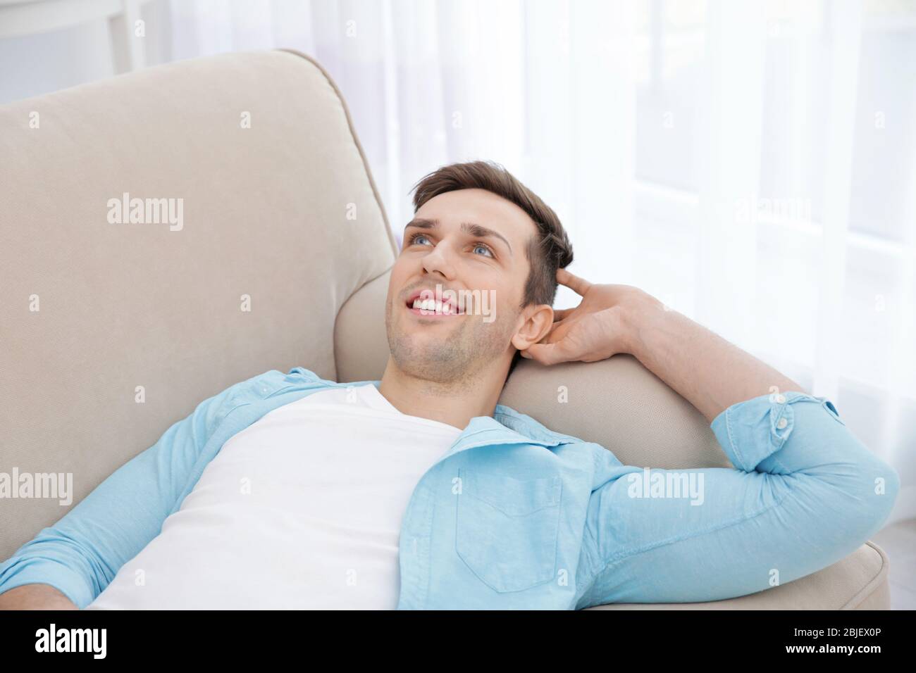 Young man resting on couch with hand behind his head in light room ...