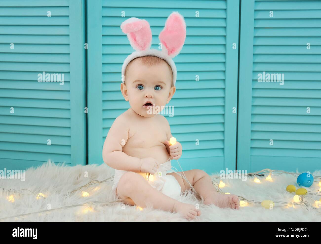 Cute little baby wearing bunny ears sitting on furry rug with garland ...