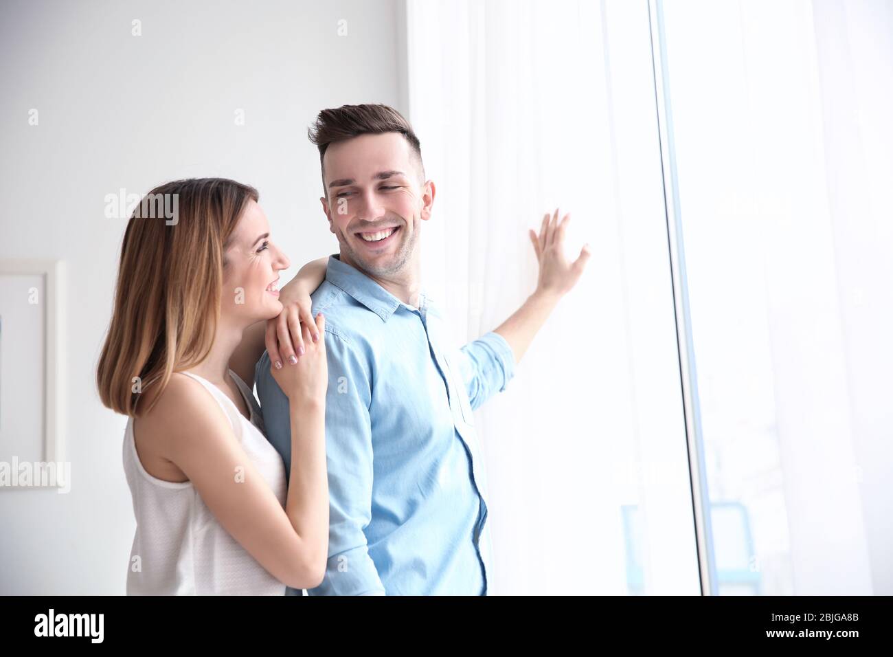 Happy couple standing near window and opening curtains Stock Photo - Alamy