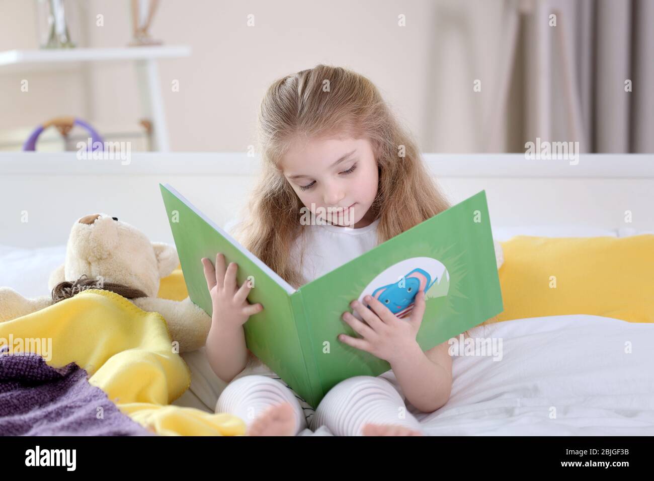Small girl sitting on bed and reading book in light room Stock Photo ...