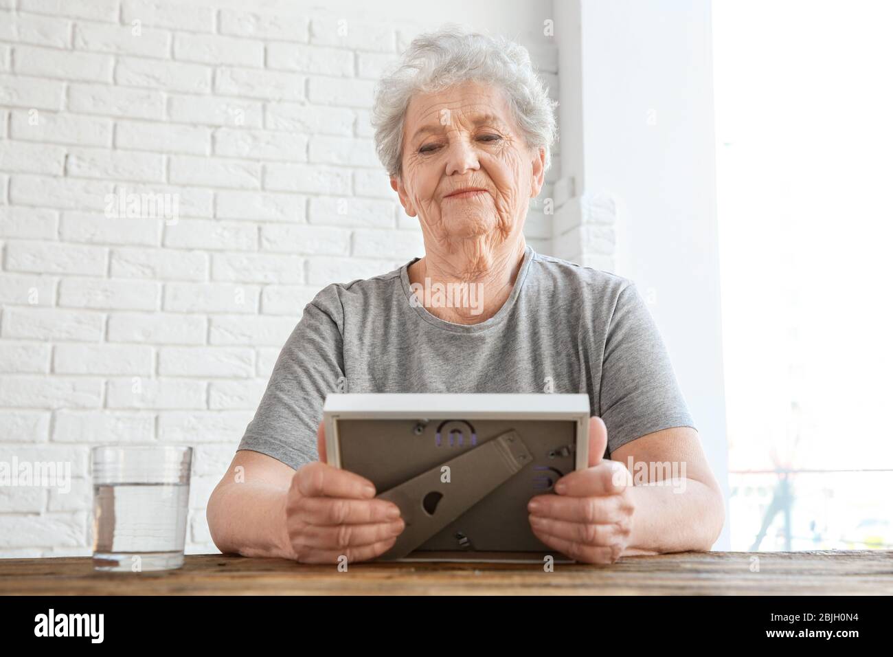 Elderly woman sitting at table with photo frame and glass of water ...