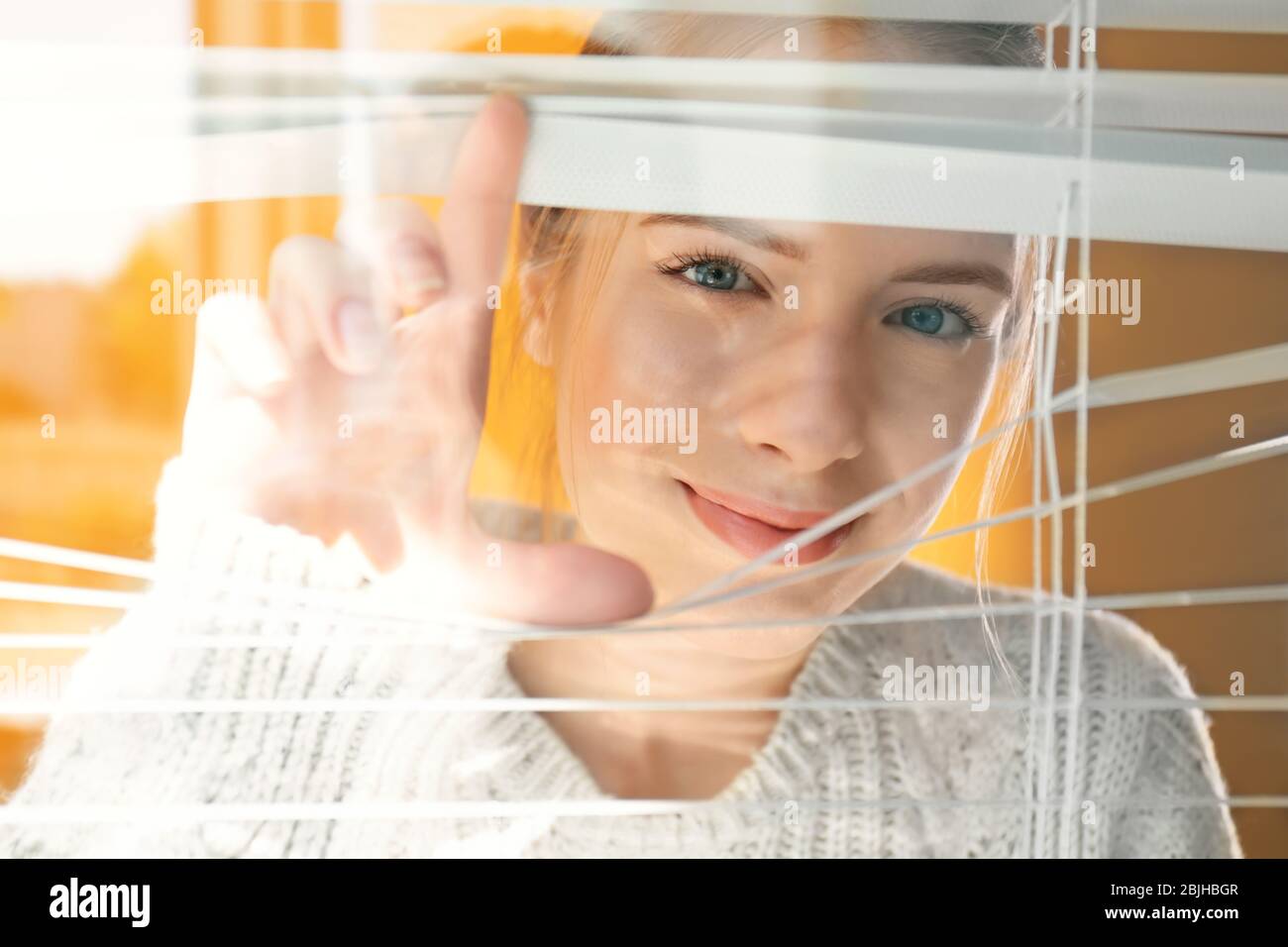 Beautiful young girl separating slats of blinds and looking through ...