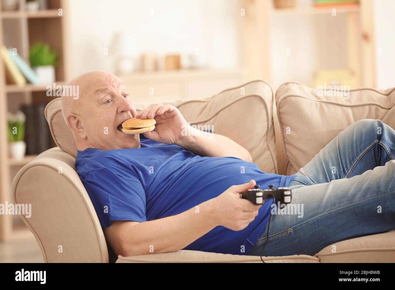 Fat senior man playing videogame and eating hamburger while lying on ...