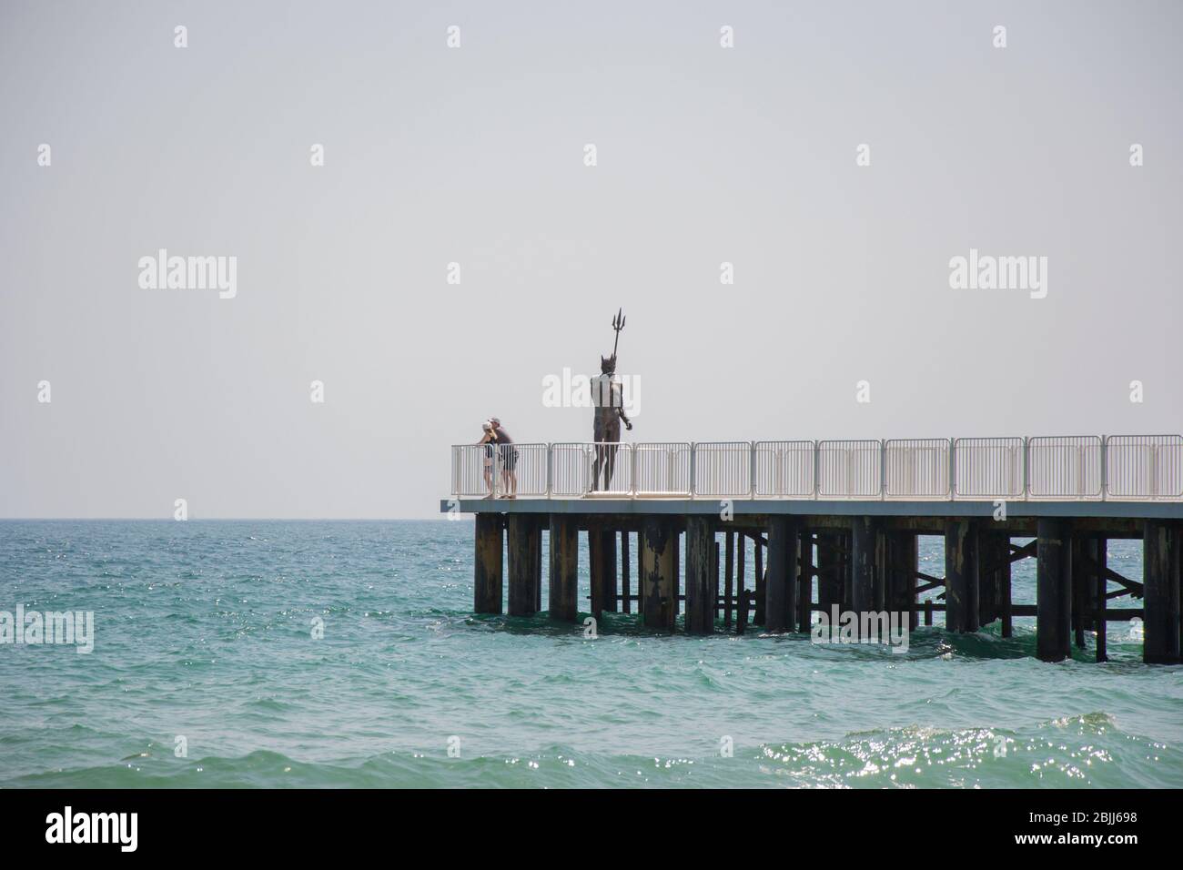 People walking on a pier with statue of Poseidon, bridge near the beach ...