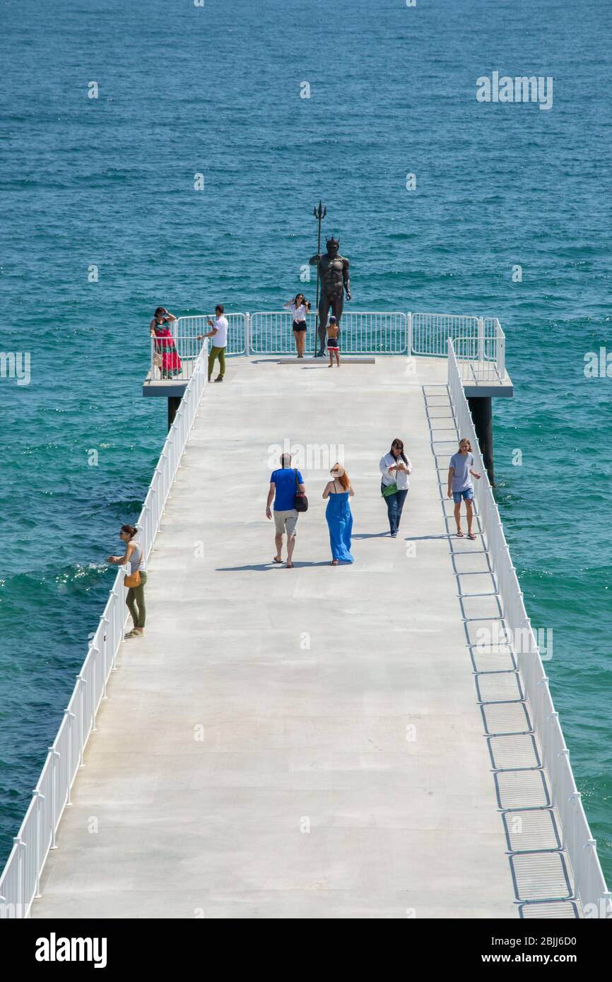 People walking on a pier with statue of Poseidon, bridge near the beach ...