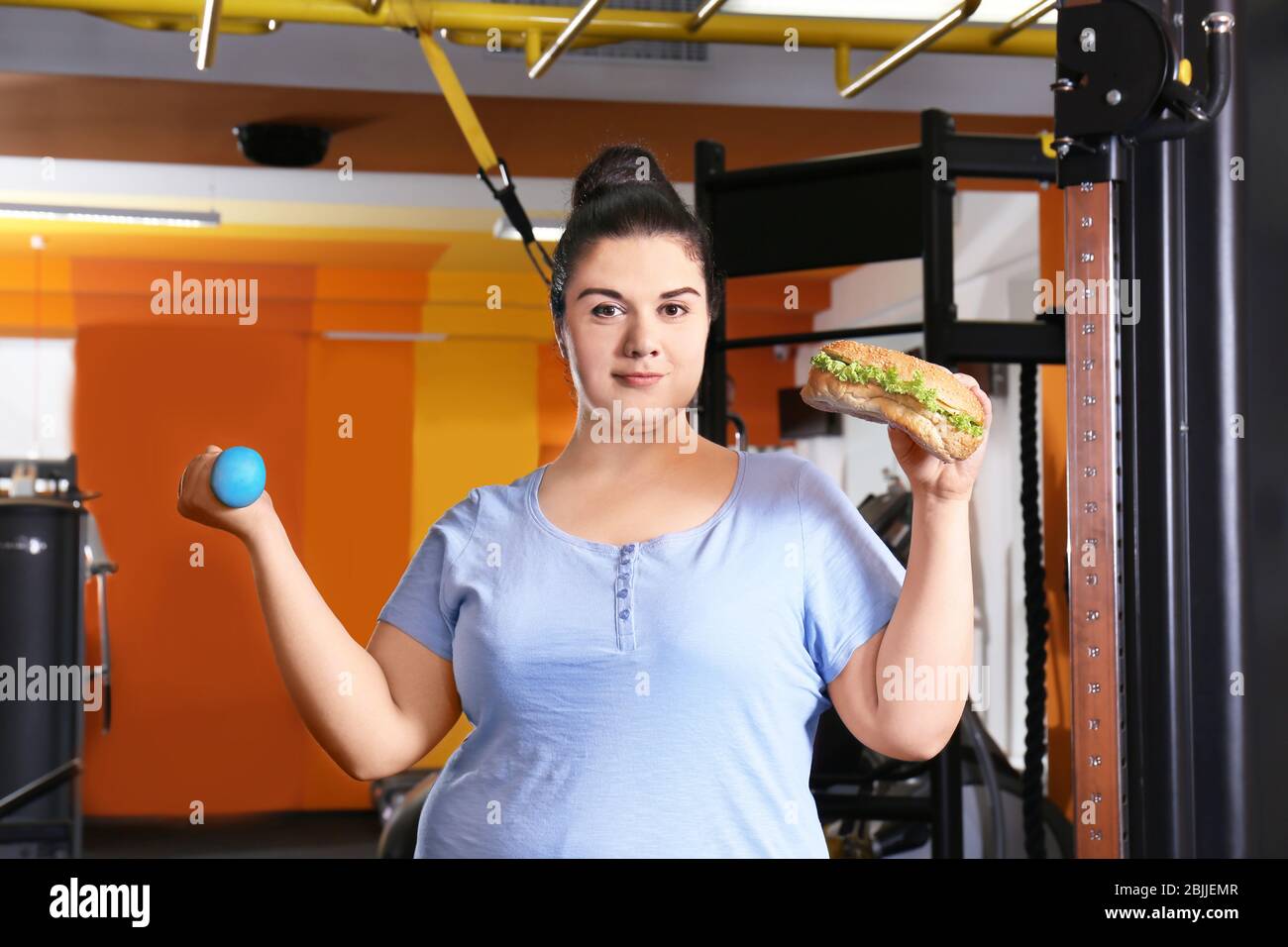 Overweight young woman eating sandwich and holding dumbbell in gym ...