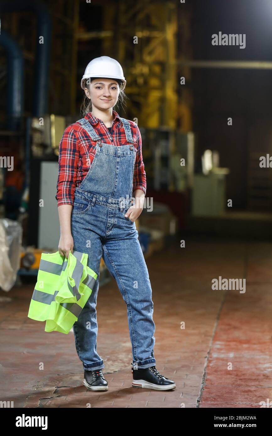 Young girl in a work dress and white hard hat in a factory. Woman in a ...