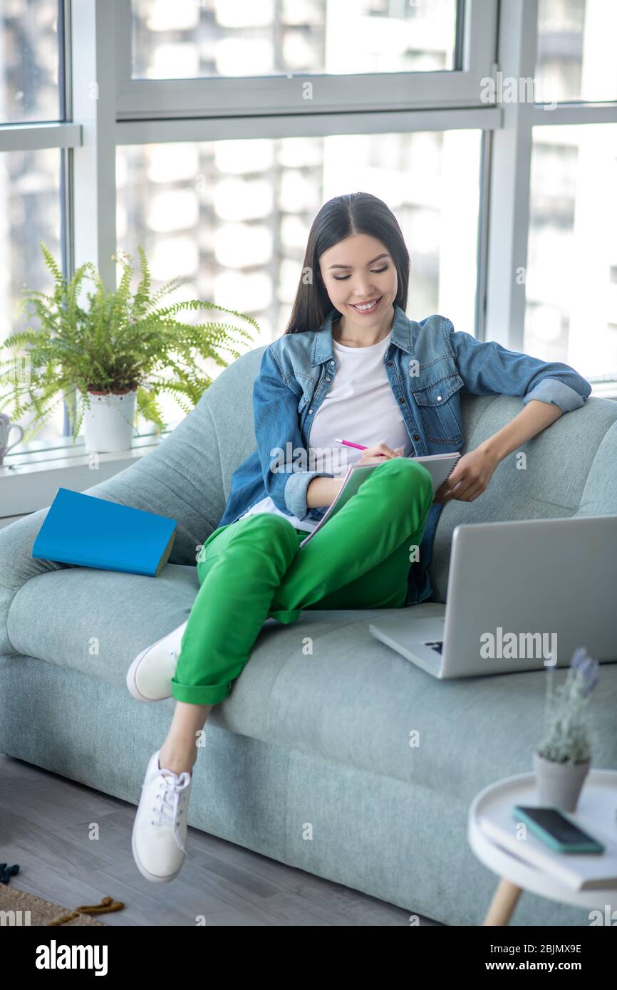 Young woman in a jeans jacket sitting on the sofa and writing something ...