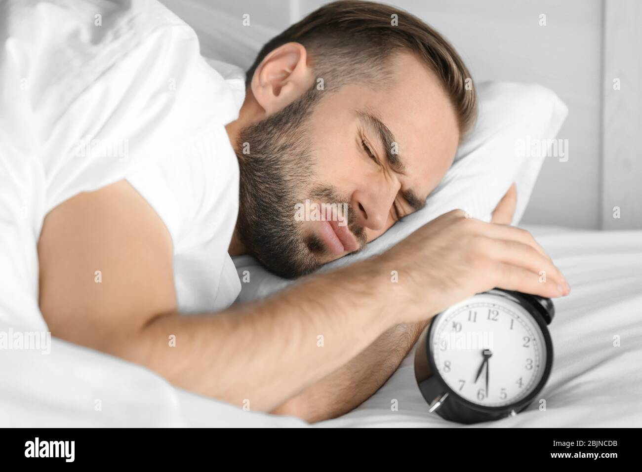 Young Man Switching Off Alarm Clock Signal While Lying In Bed At Home