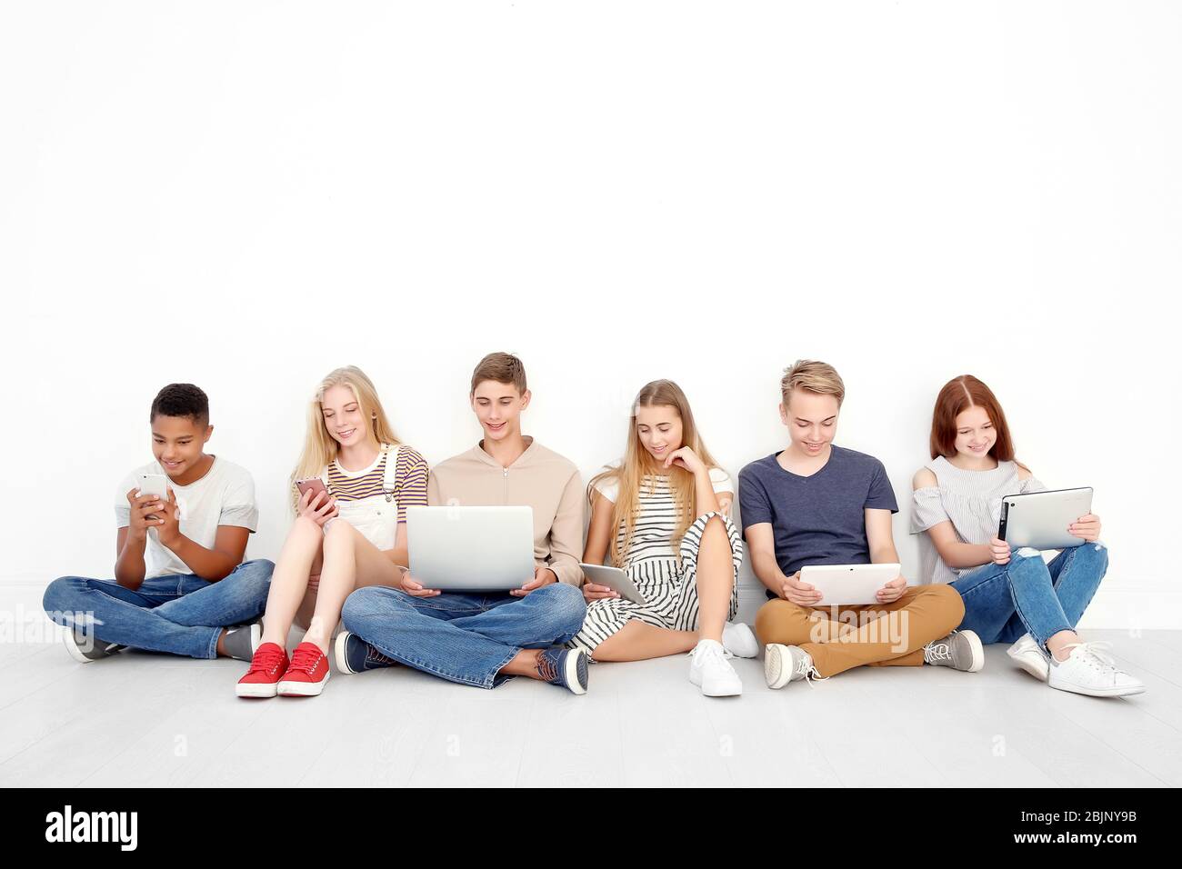 Teenagers with modern gadgets sitting on floor near light wall Stock ...