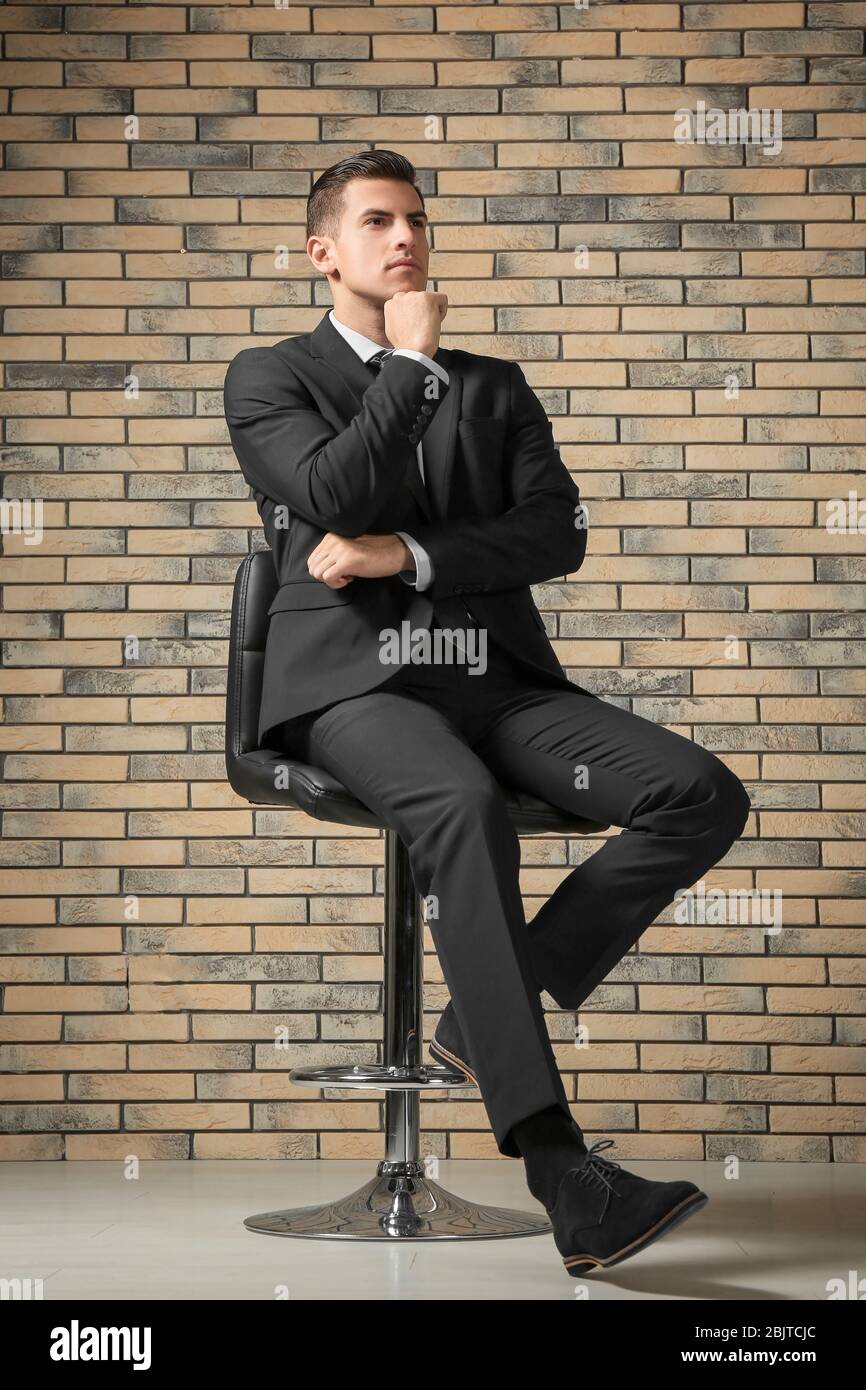 Handsome man in elegant suit sitting on chair near brick wall Stock ...