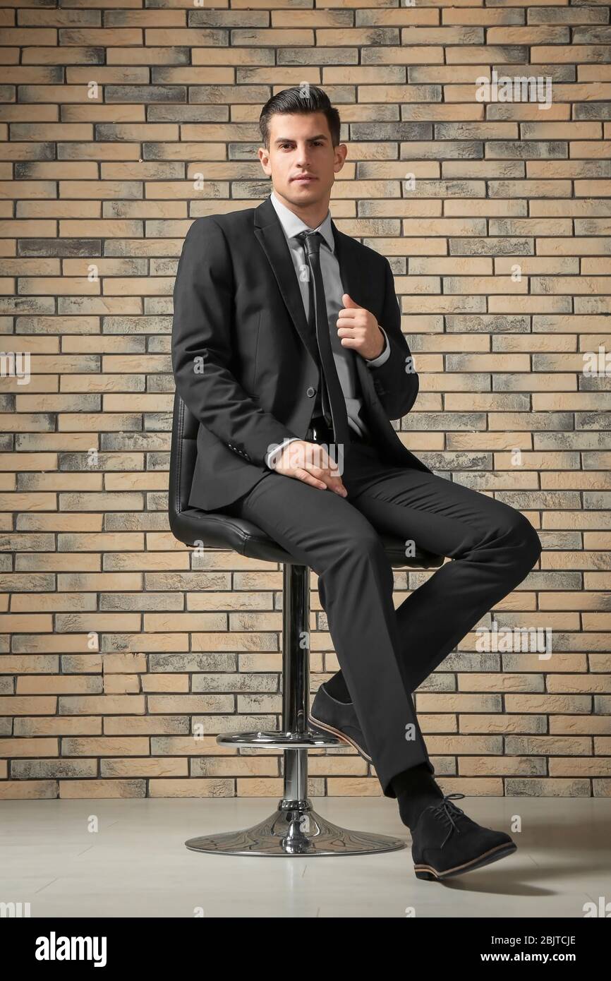 Handsome man in elegant suit sitting on chair near brick wall Stock ...