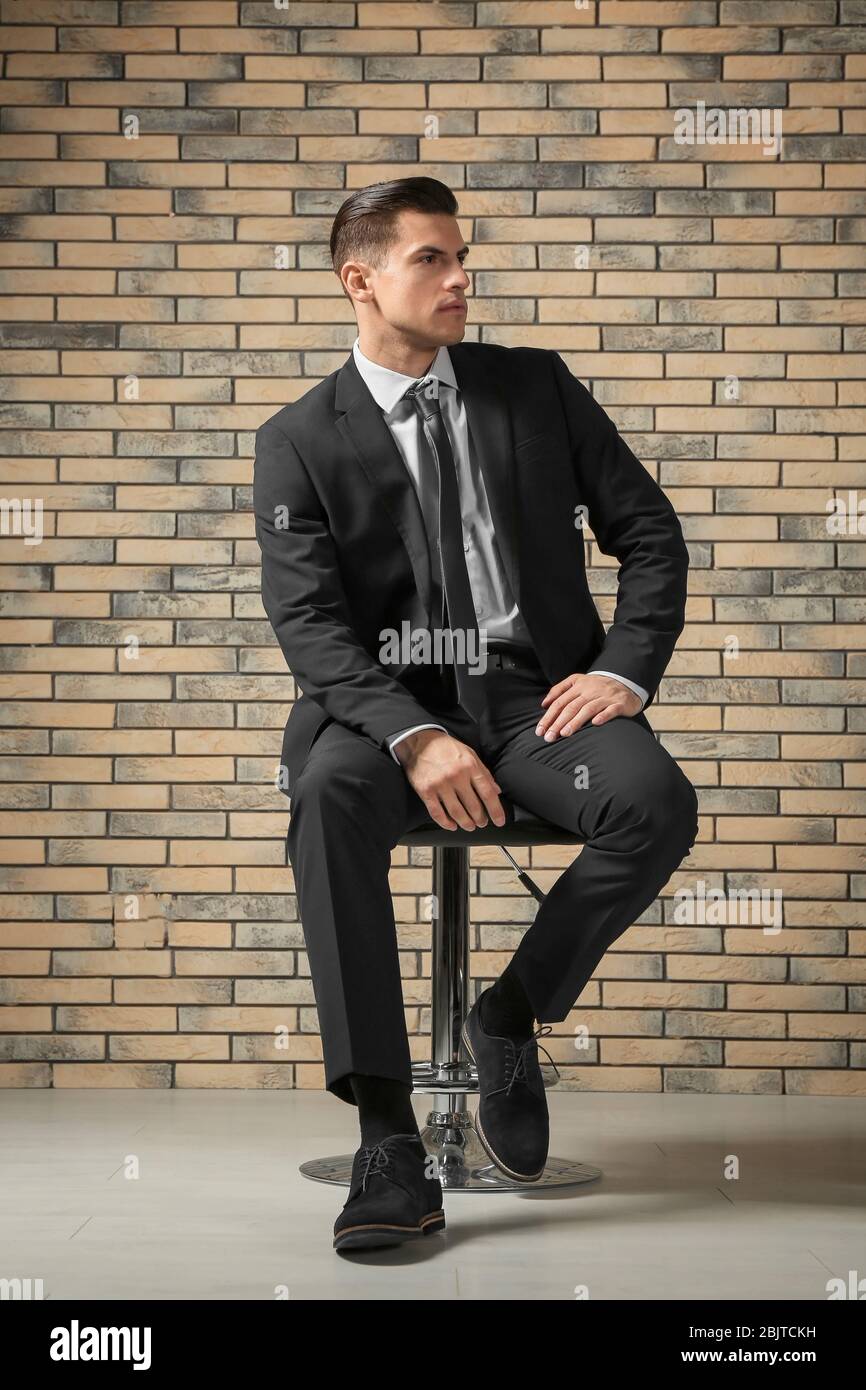 Handsome man in elegant suit sitting on chair near brick wall Stock ...