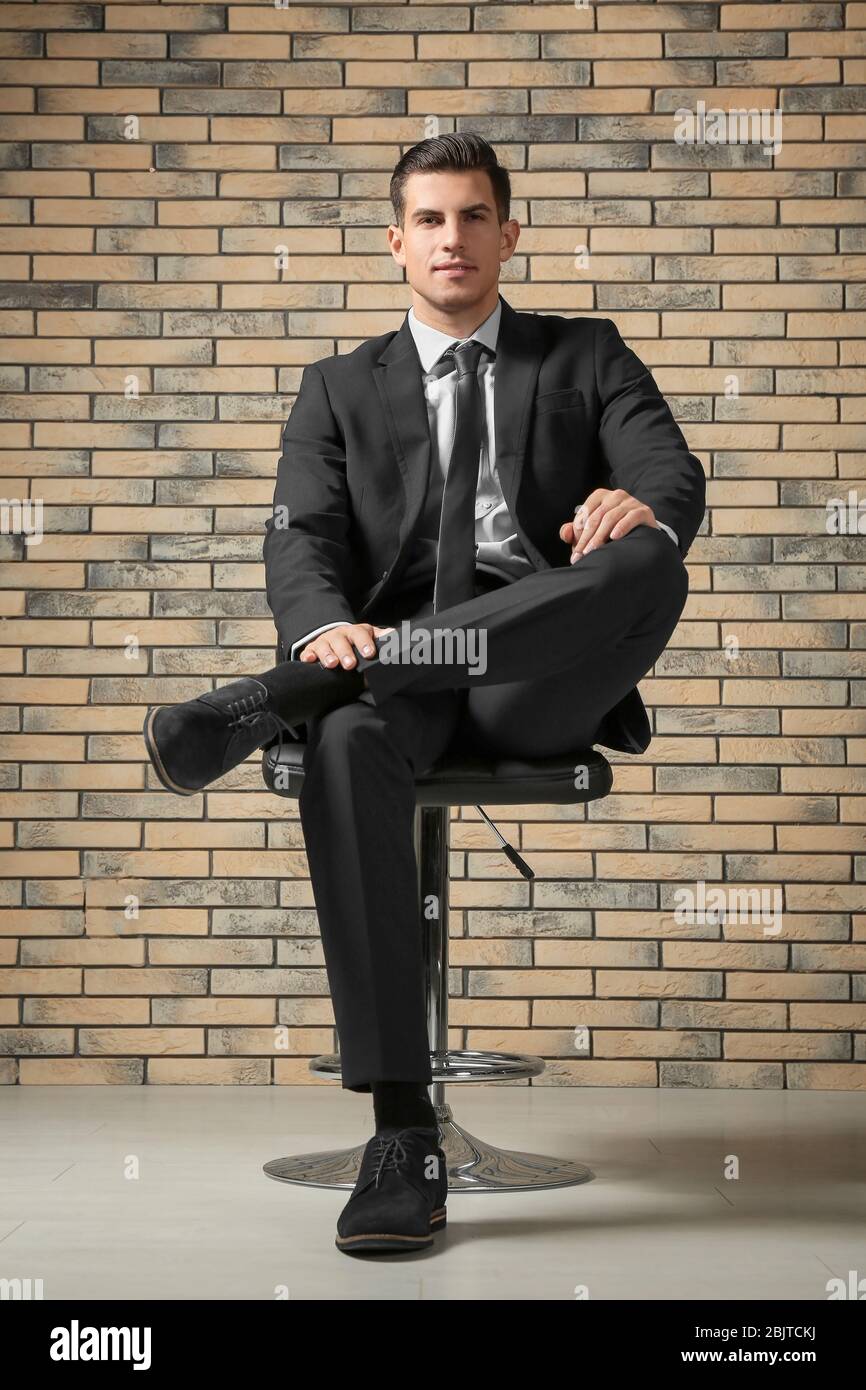 Handsome man in elegant suit sitting on chair near brick wall Stock ...