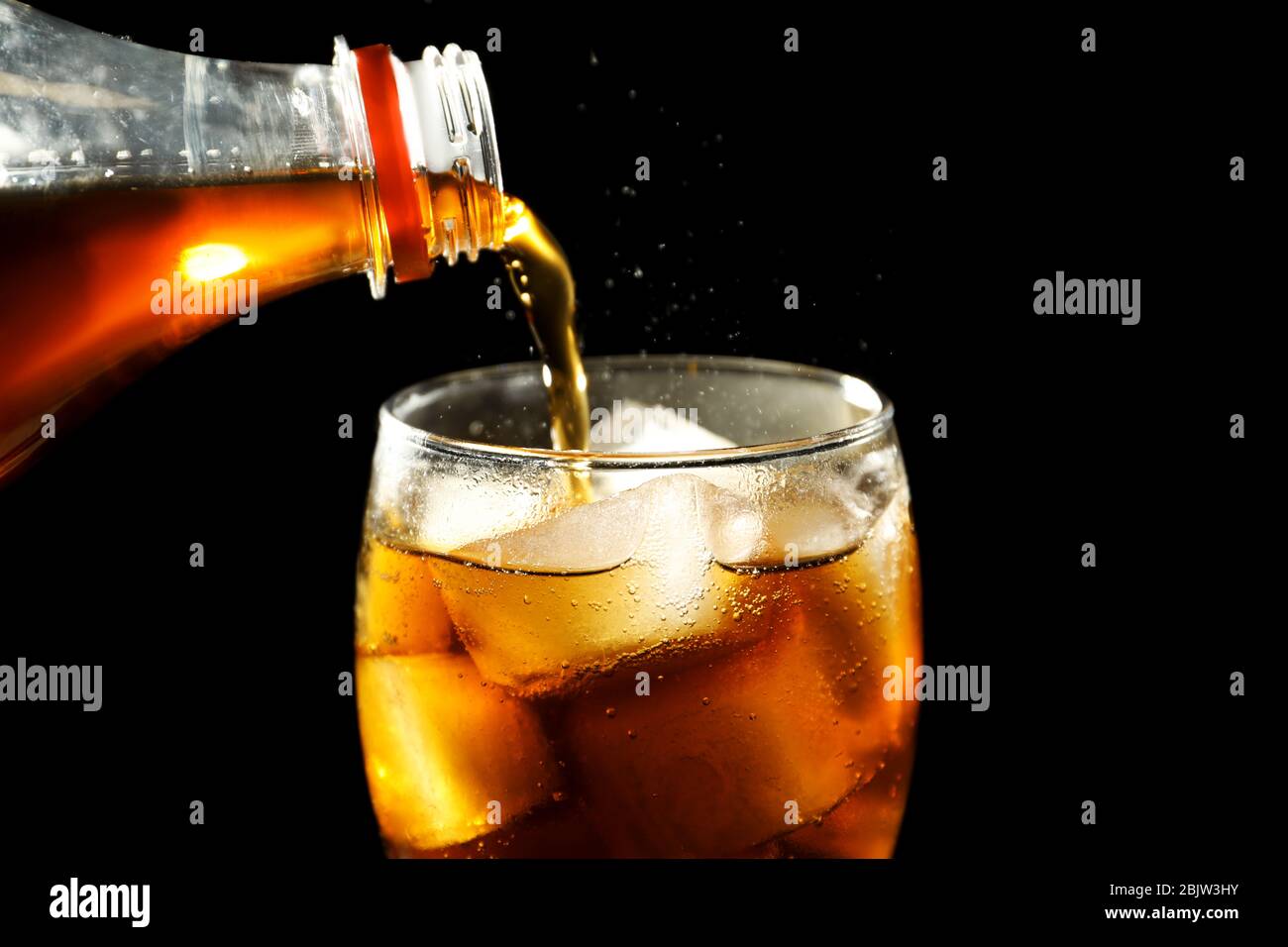 Pouring cola from bottle into glass with ice against black background ...