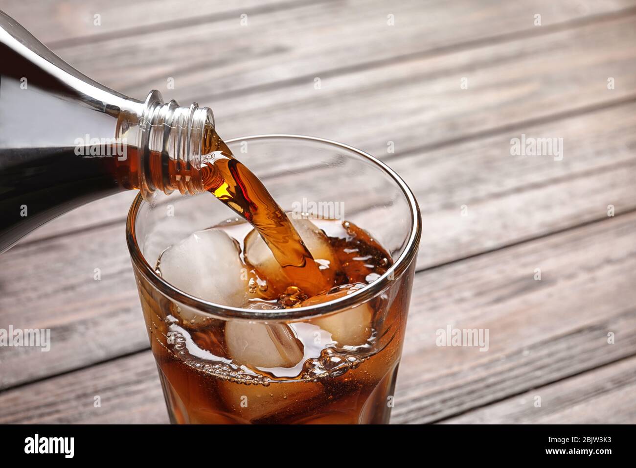 Pouring cola from bottle into glass with ice on wooden background ...