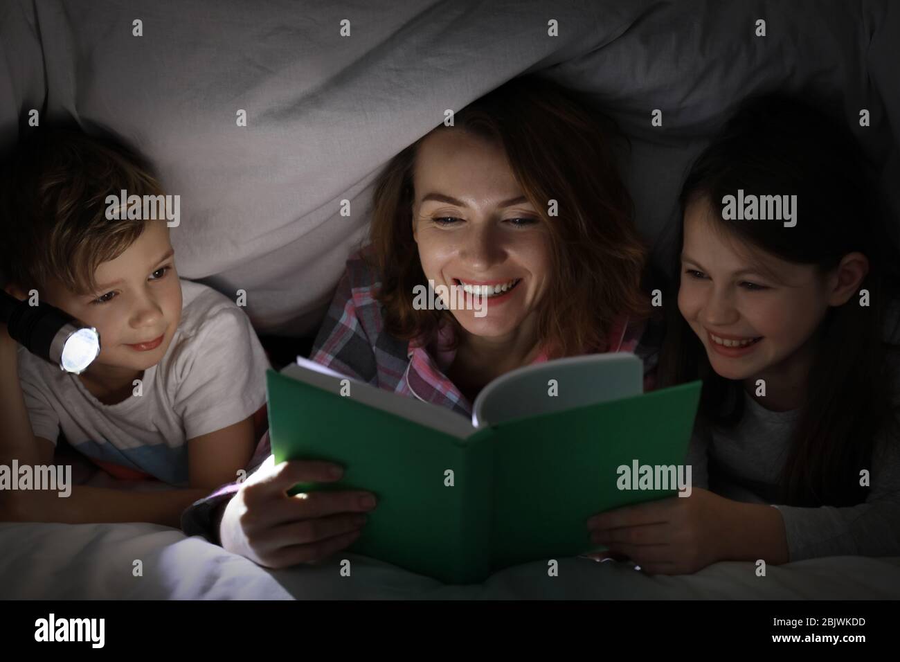 Mother reading bedtime story to her little children under blanket in ...
