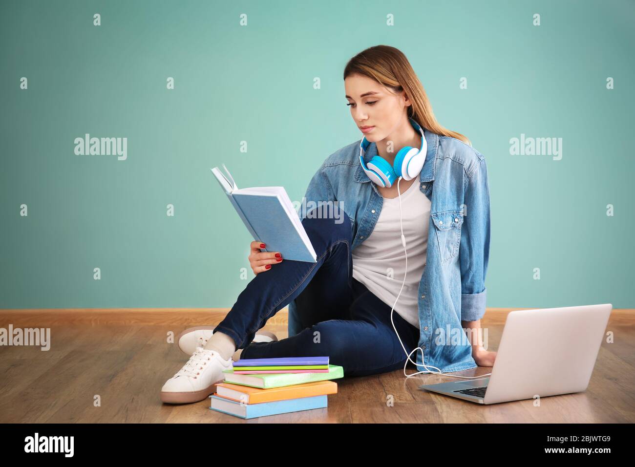 Pretty student with laptop and book studying on floor against color ...