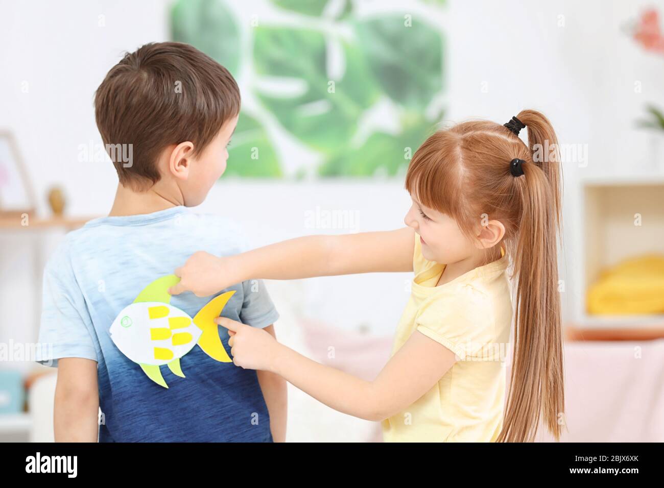 Little girl sticking paper fish to boy's back indoors. April fool's day ...