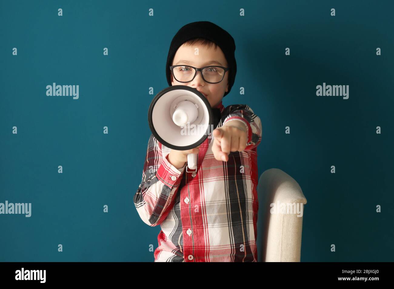 Cute little boy with megaphone sitting on chair against color ...