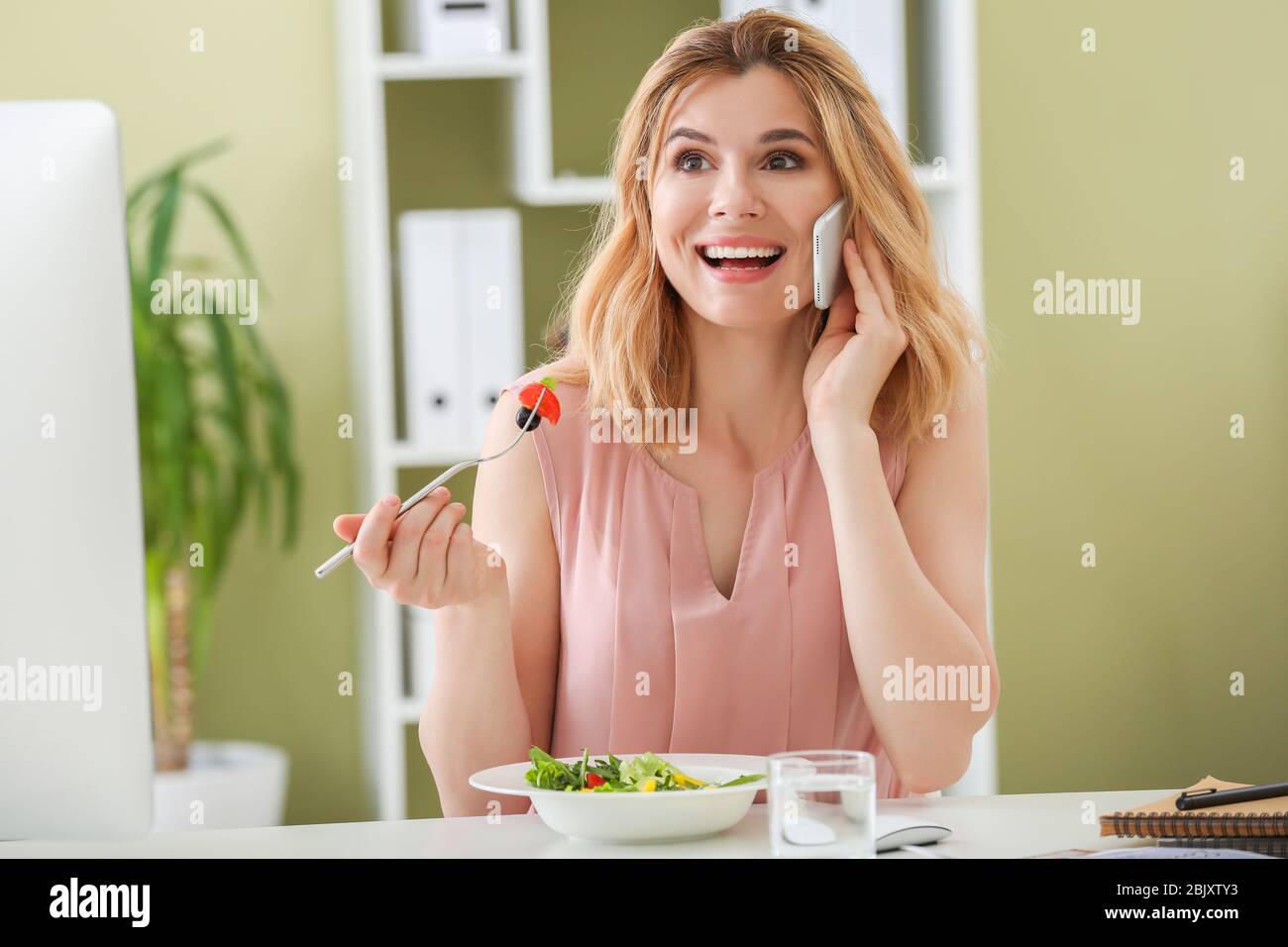 Woman eating healthy vegetable salad while talking by phone in office ...