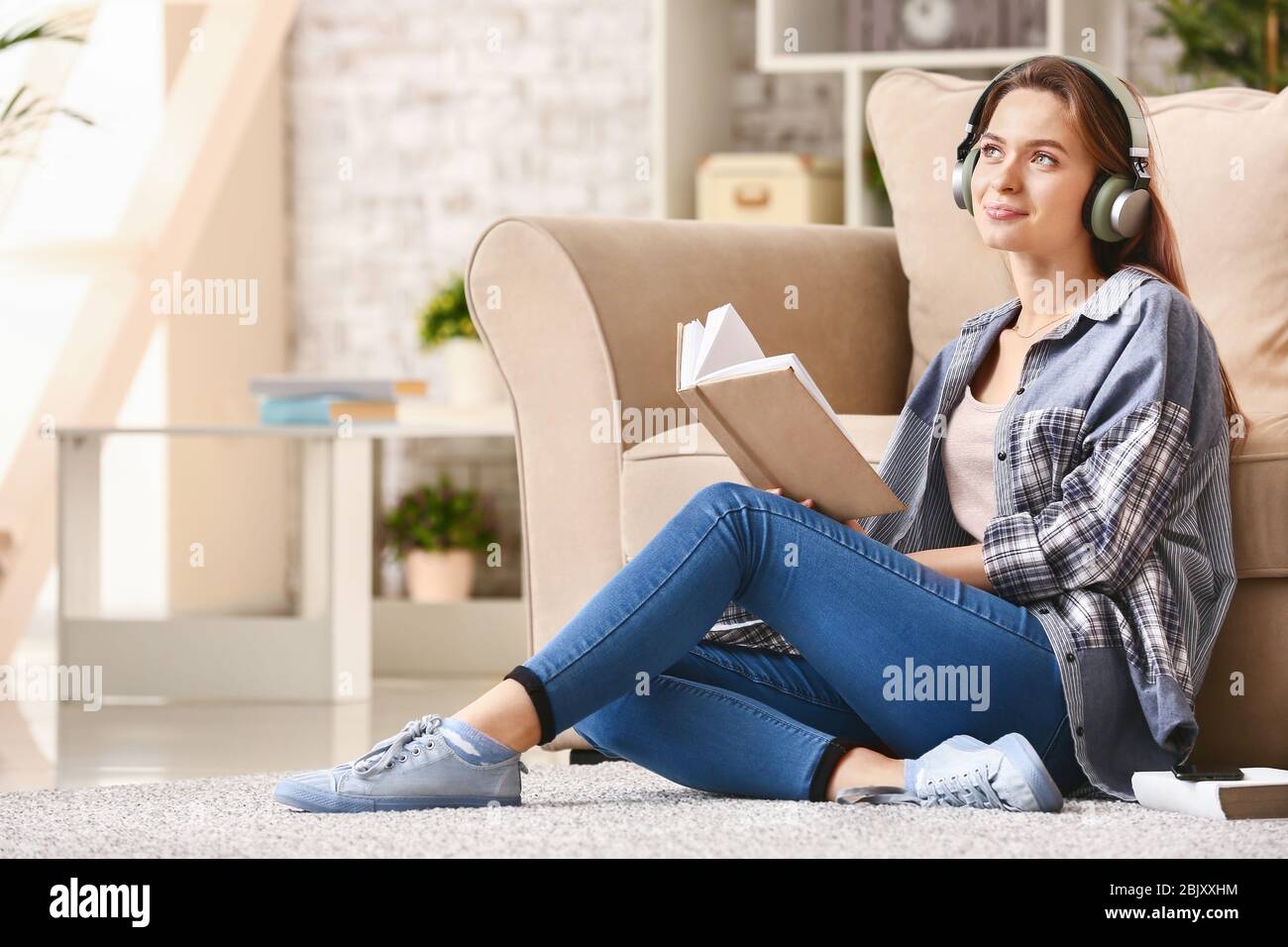 Beautiful young woman listening to music while reading book at home ...