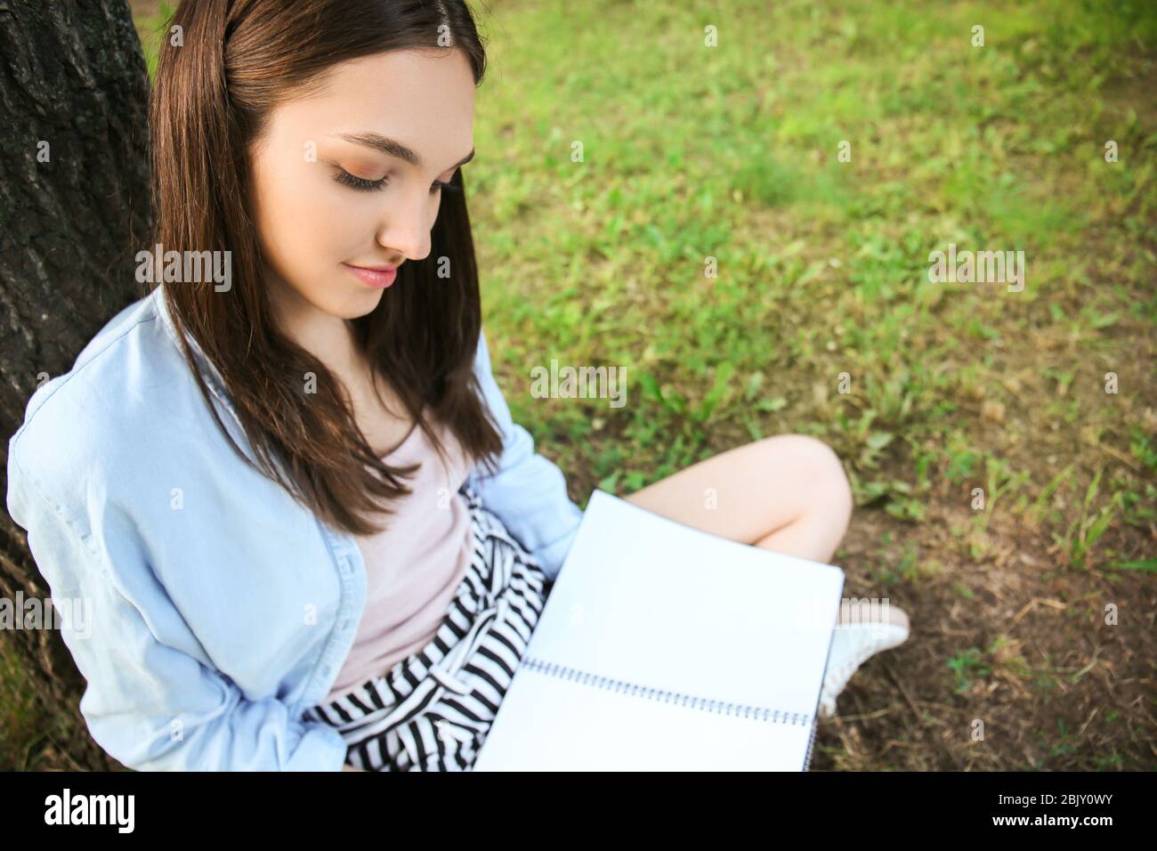 Beautiful female student sitting under tree in park Stock Photo - Alamy