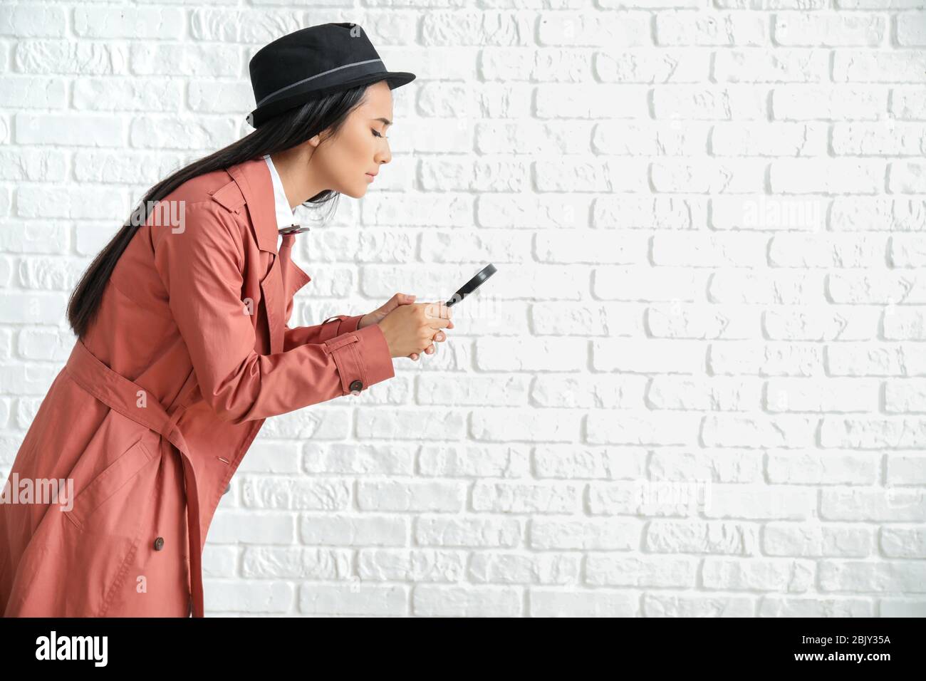 Female Asian detective with magnifying glass against white brick wall ...