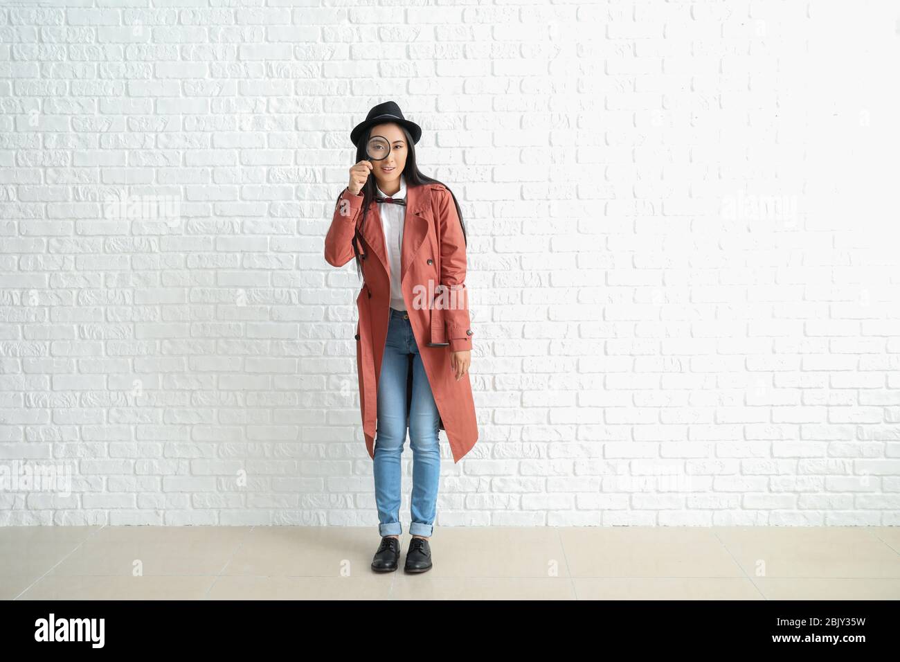 Female Asian detective with magnifying glass against white brick wall ...