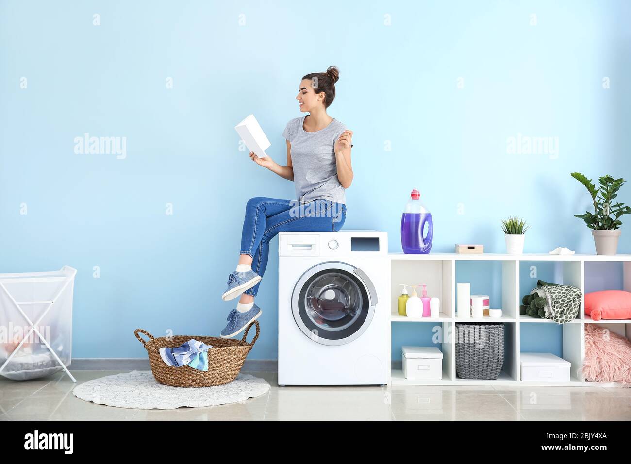 Beautiful young woman reading book while doing laundry at home Stock ...