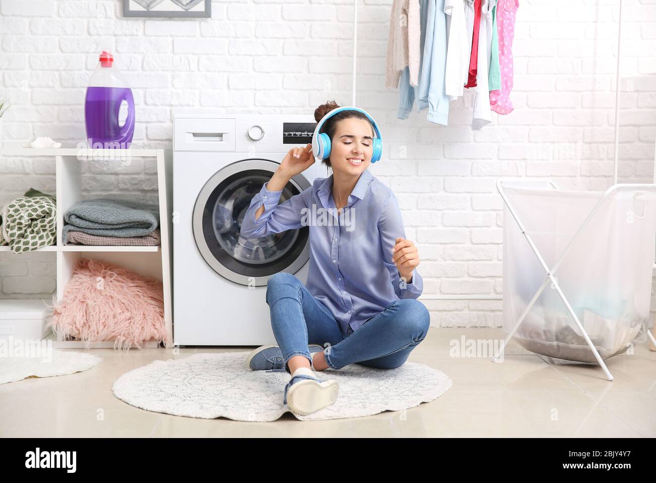 Beautiful young woman listening to music while doing laundry at home ...