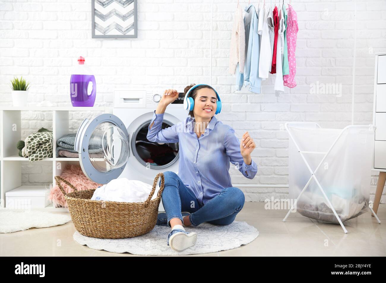 Beautiful young woman listening to music while doing laundry at home ...