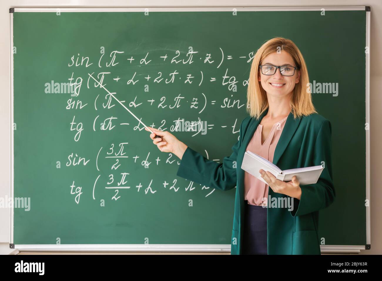 Beautiful math teacher with book and pointer near blackboard in ...