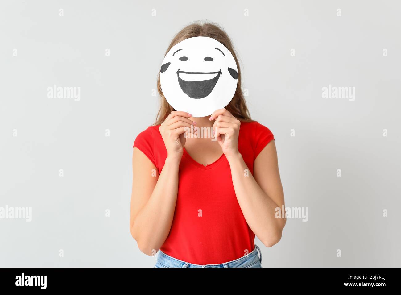 Young woman hiding face behind emoticon against light background Stock ...