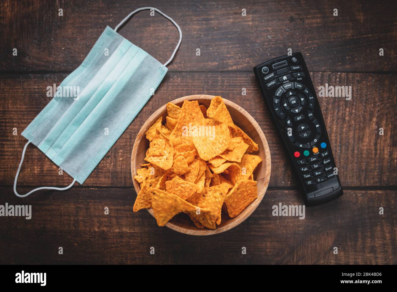 Remote control, bowl of crisps and medical mask on wooden table ...