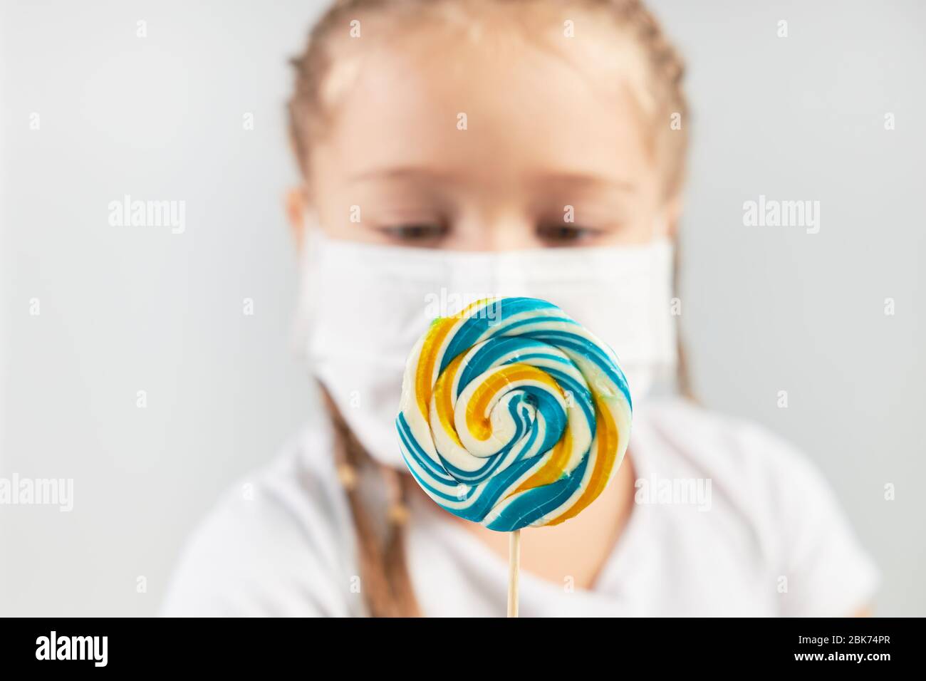 Child in a white medical mask looks at a delicious lollipop. Focus on ...