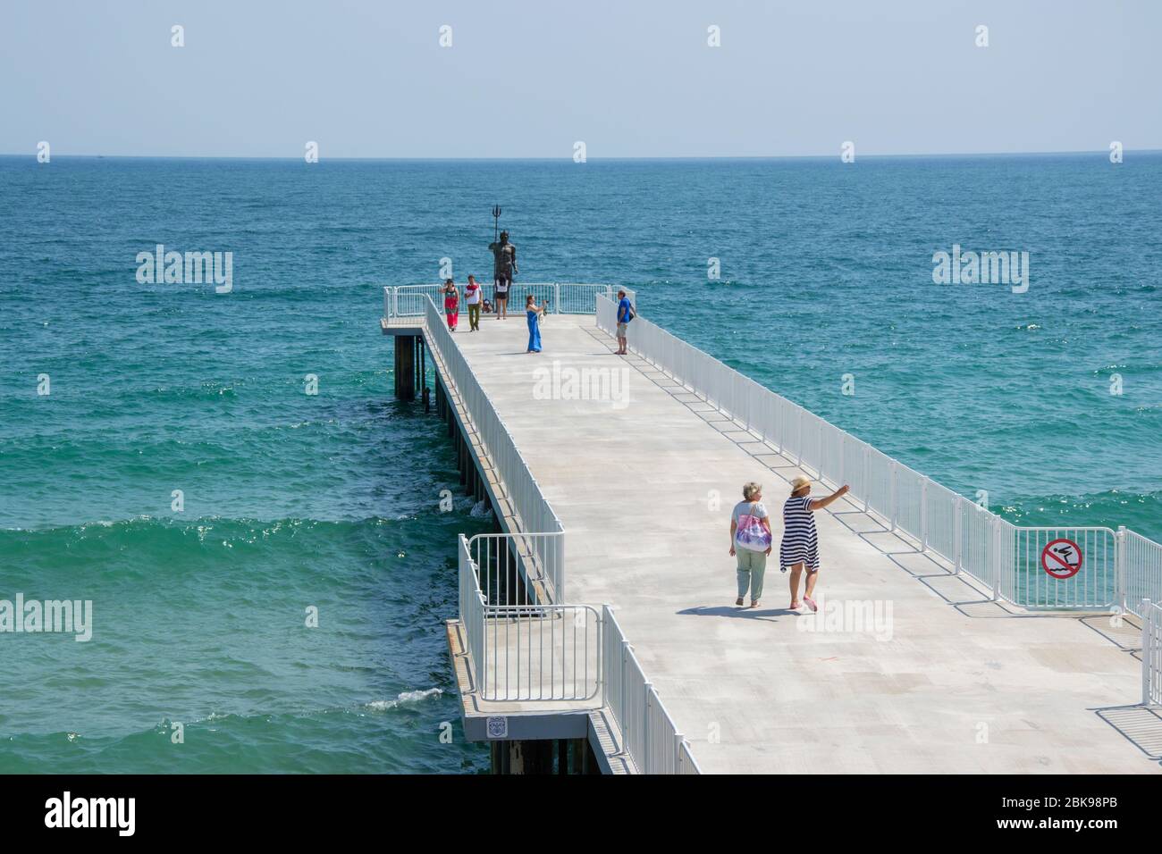 People walking on a pier with statue of Poseidon, bridge near the beach ...