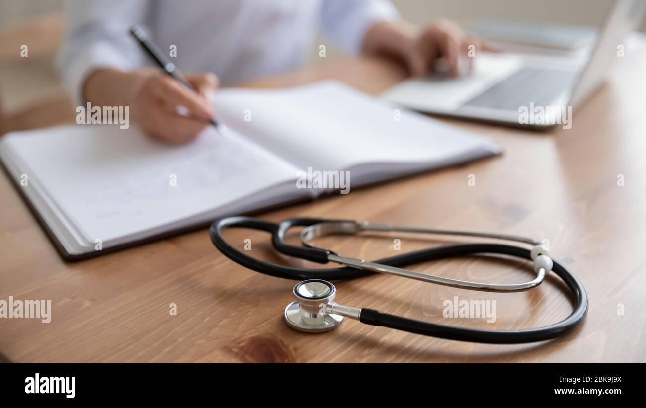 Female doctor using laptop, writing notes with stethoscope on table ...