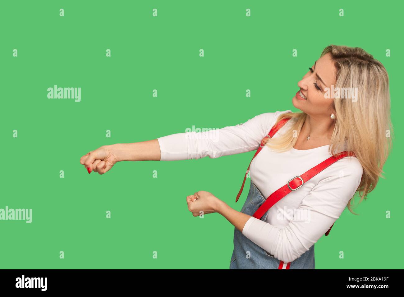 Portrait of persistent adult woman in denim overalls pulling invisible ...