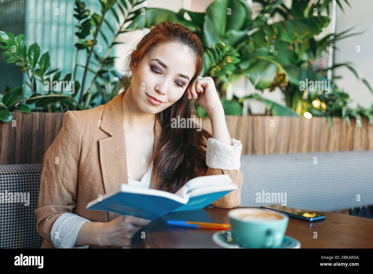 Young brunette asian girl student reading book with coffee on the table ...