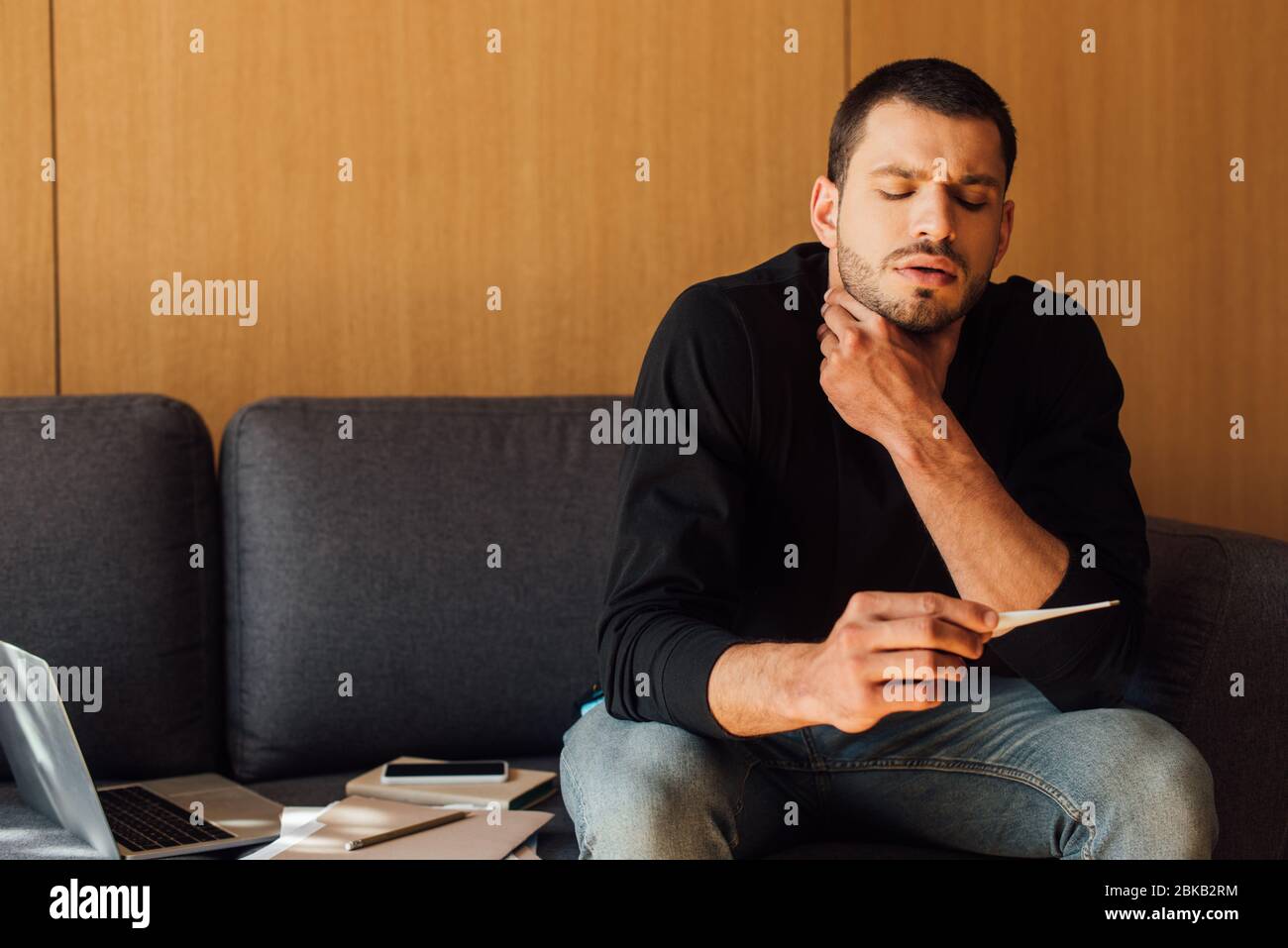 sick man with sore throat looking at digital thermometer near laptop ...