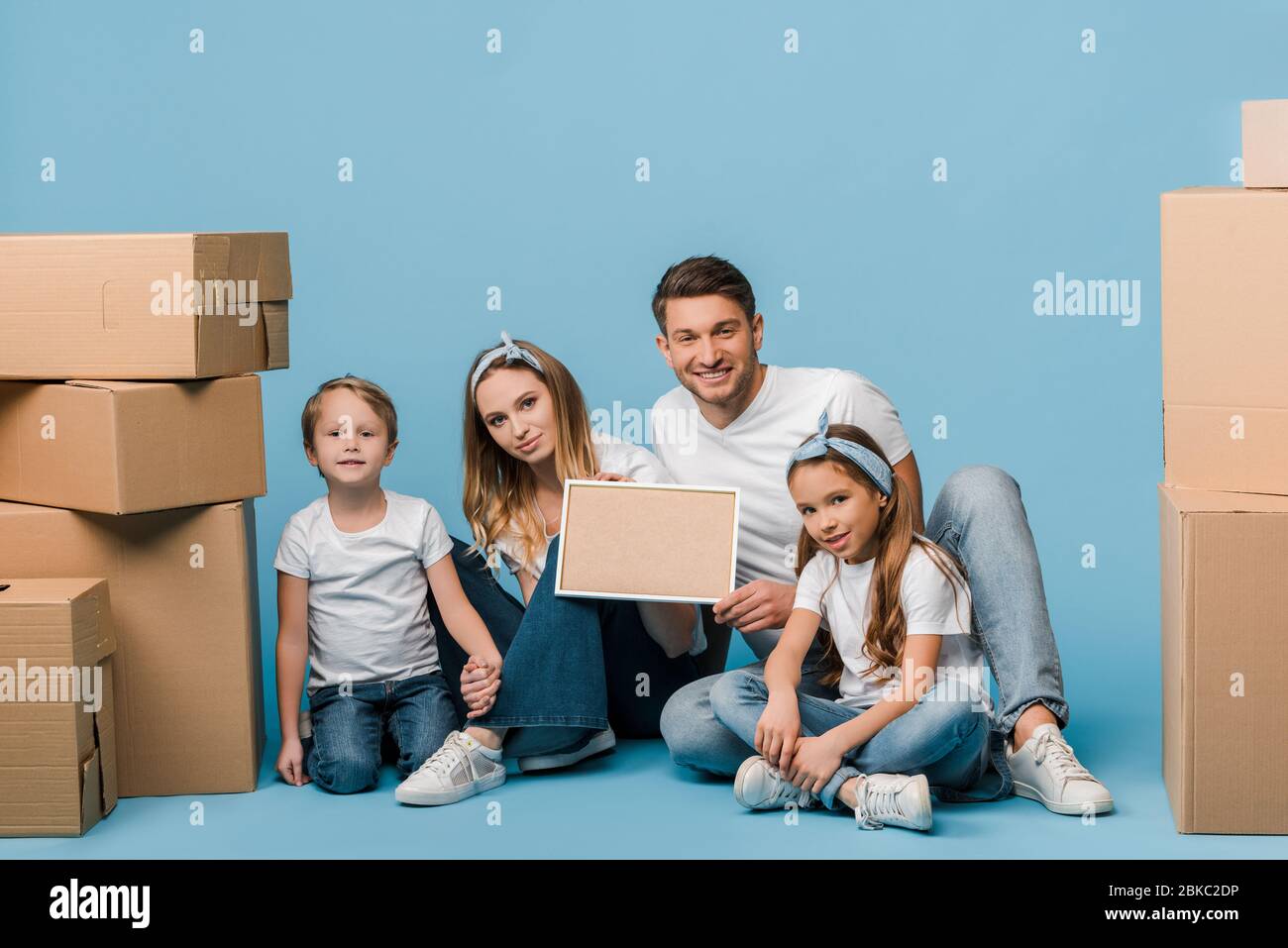happy parents and kids holding frame and sitting on blue with cardboard ...
