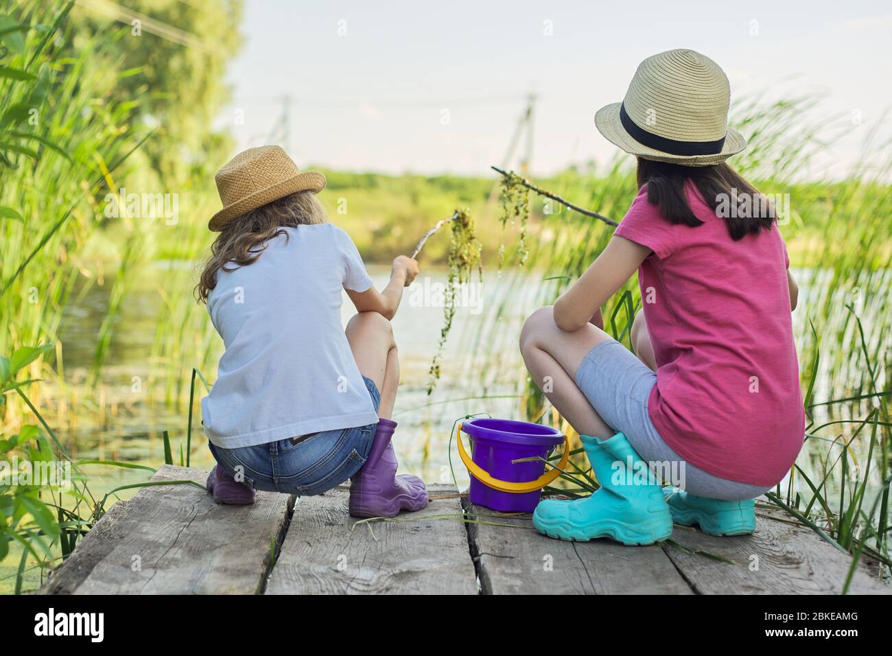 Kids girls sitting on wooden pier, catching water snails in bucket ...