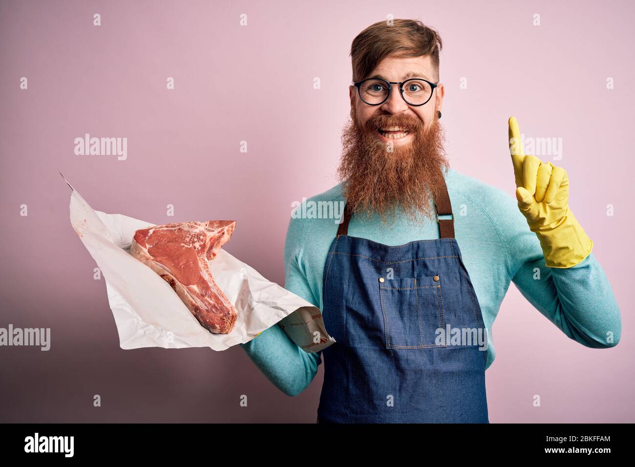 Redhead Irish butcher man with beard holding raw beef steak over pink ...